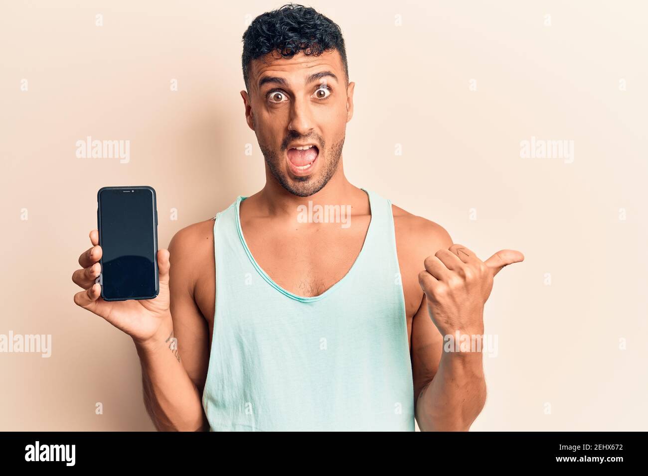 Young hispanic man holding smartphone showing screen pointing thumb up to the side smiling happy ...
