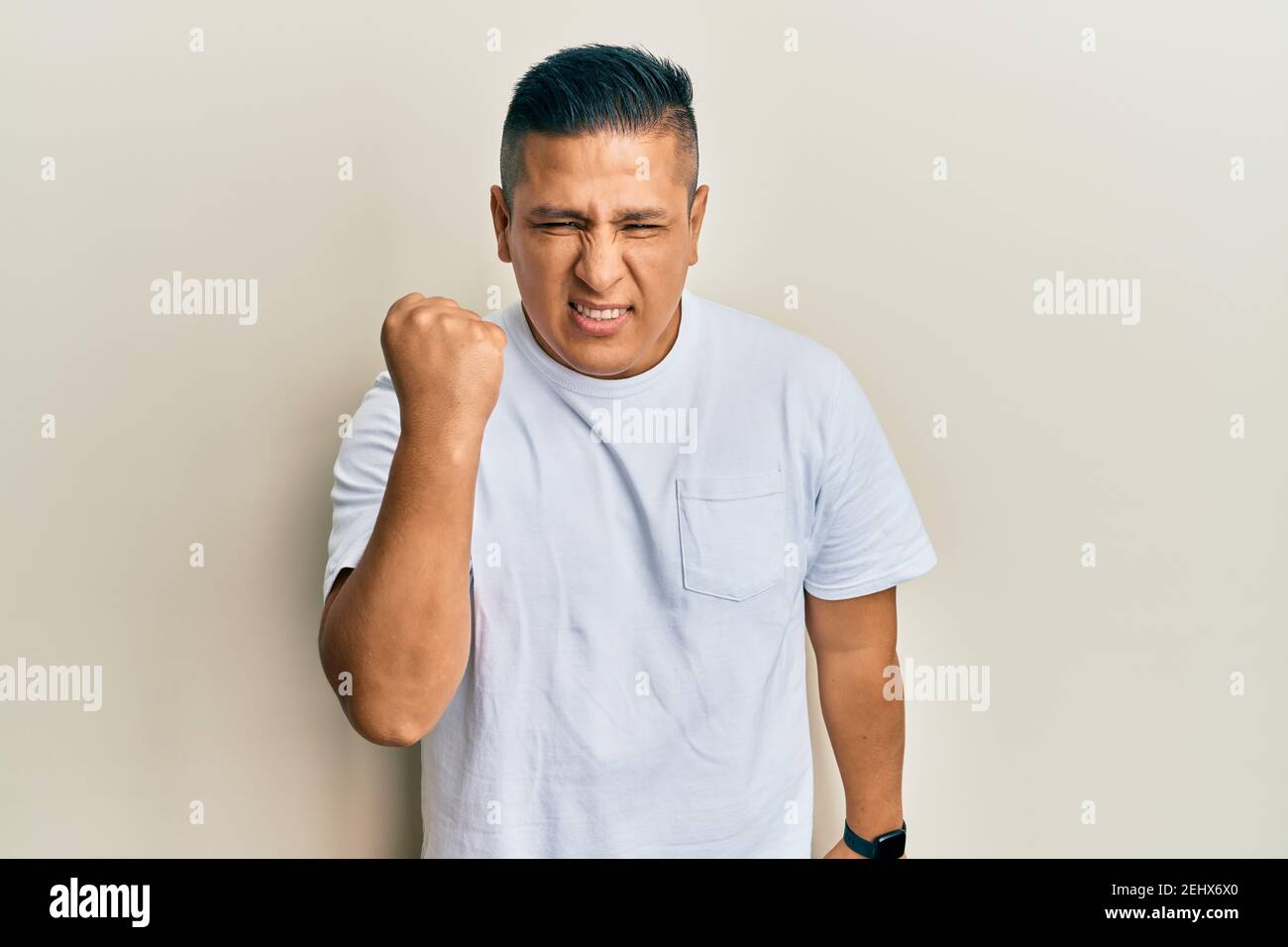 Young latin man wearing casual white t shirt angry and mad raising fist ...