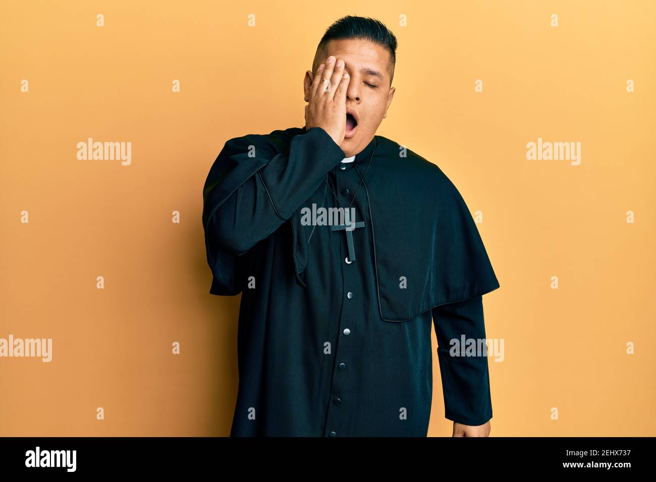 Young latin priest man standing over yellow background yawning tired ...