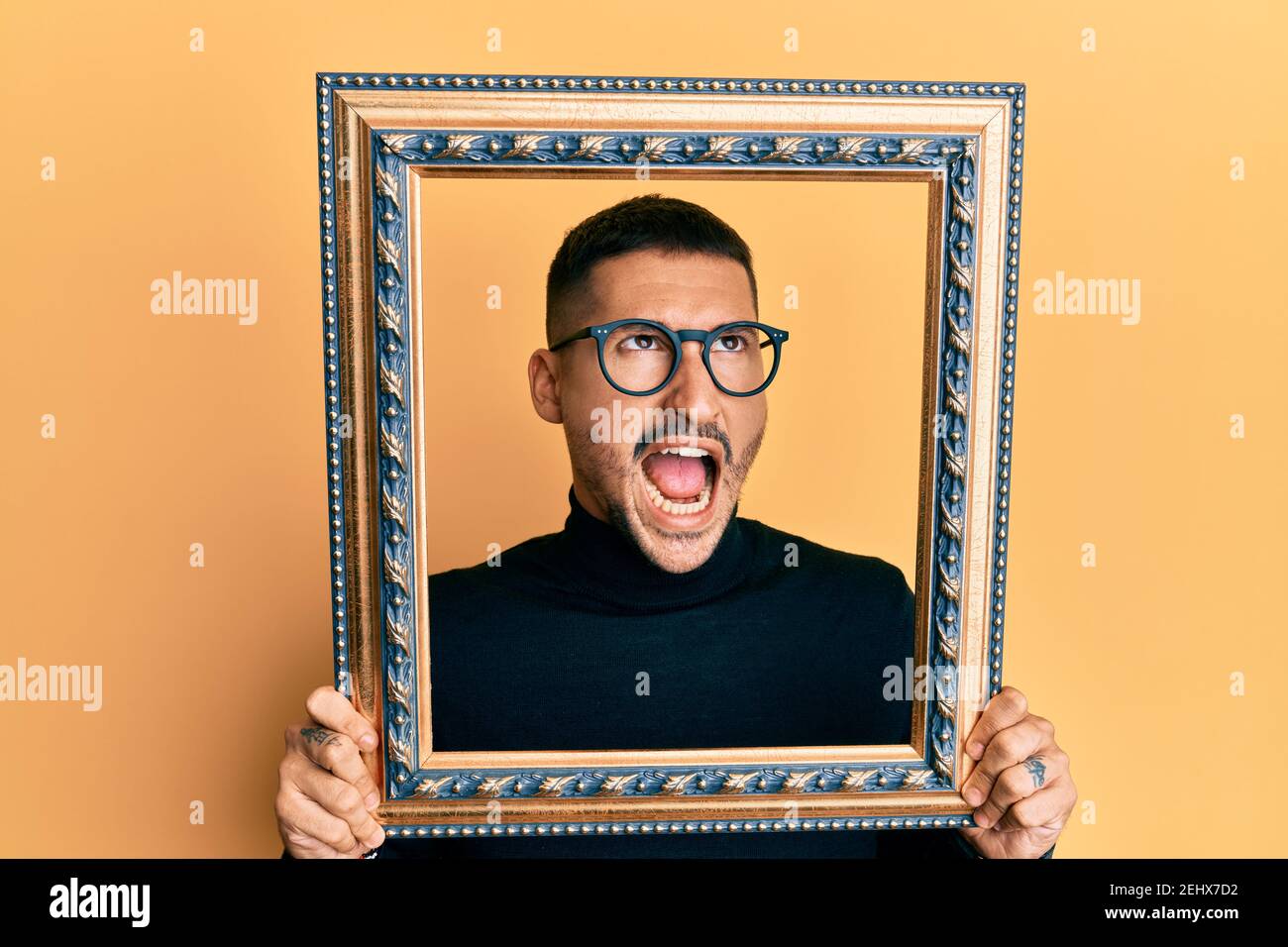 Handsome man with tattoos holding empty frame angry and mad screaming ...