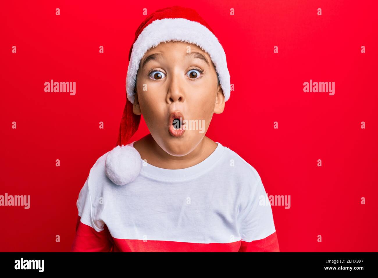 Little boy hispanic kid wearing christmas hat scared and amazed with open mouth for surprise ...