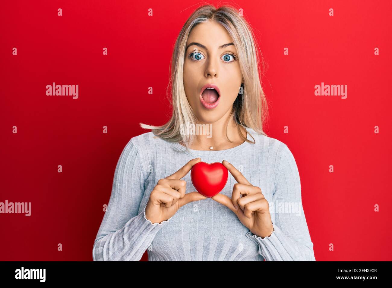 Beautiful blonde woman holding heart afraid and shocked with surprise ...