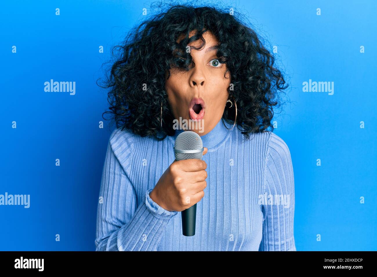 Young hispanic woman singing song using microphone scared and amazed ...