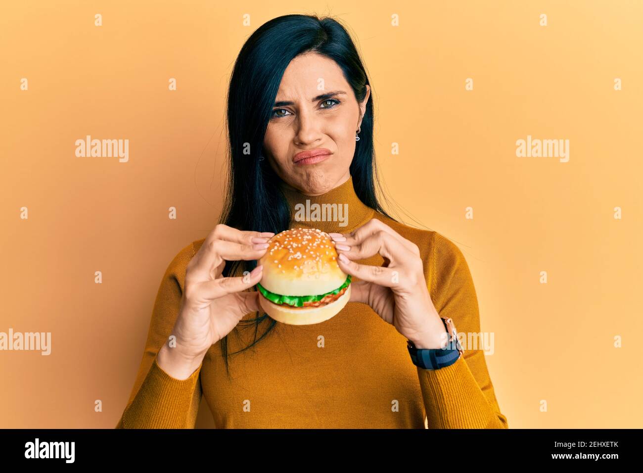 Young caucasian woman eating a tasty classic burger skeptic and nervous ...