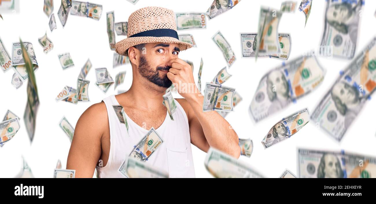 Young hispanic man wearing casual summer hat smelling something stinky ...