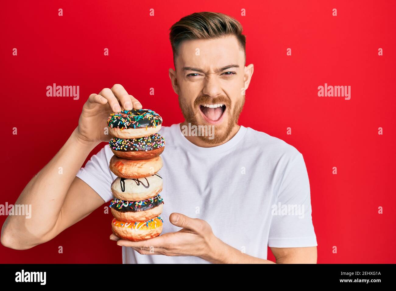 Young redhead man holding pile of tasty colorful doughnuts celebrating ...
