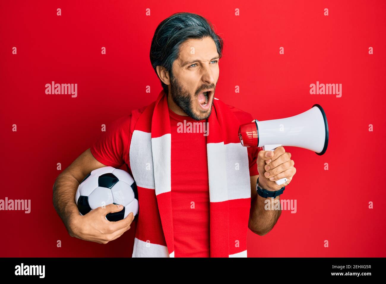 Young hispanic man football hooligan holding ball using megaphone angry ...
