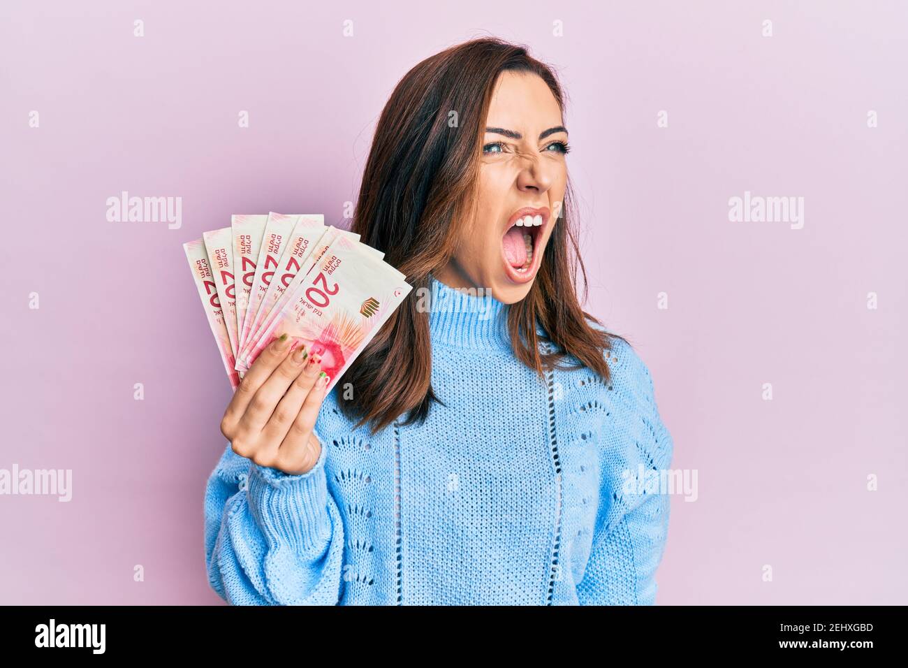 Young brunette woman holding 20 israel shekels banknotes angry and mad ...