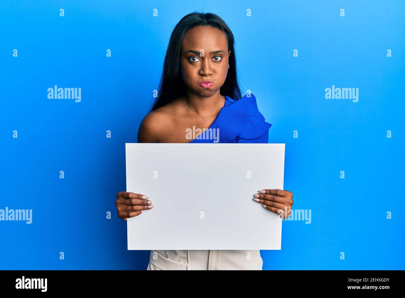 Young african woman holding blank empty banner puffing cheeks with ...