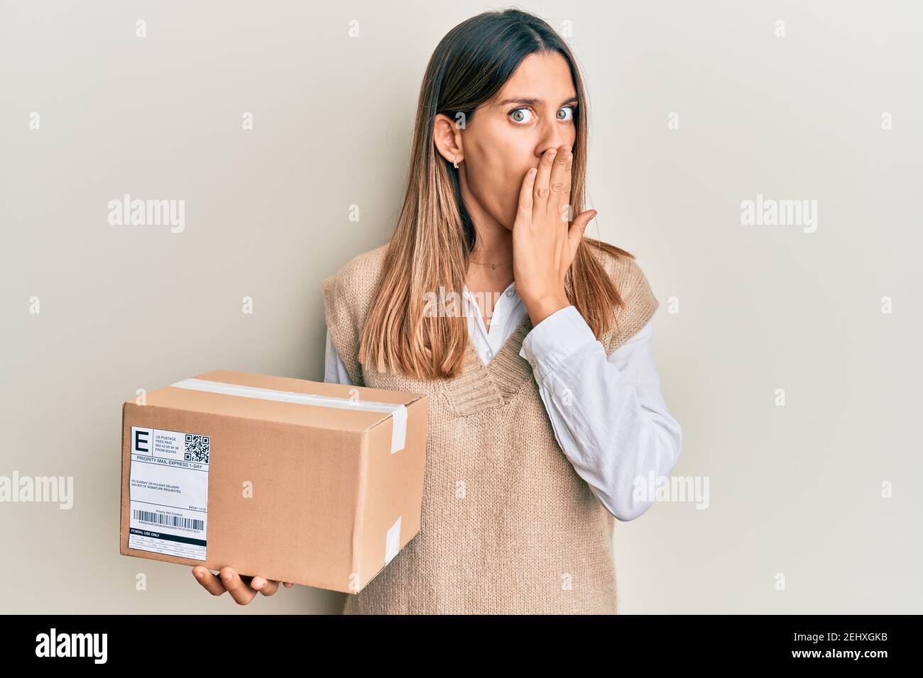 Brunette young woman holding delivery package covering mouth with hand ...