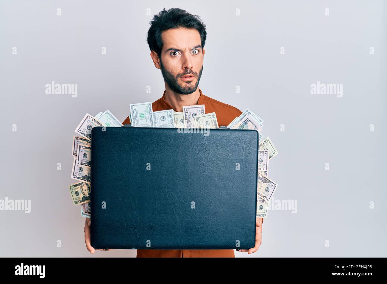 Young hispanic man holding briefcase with dollars in shock face ...