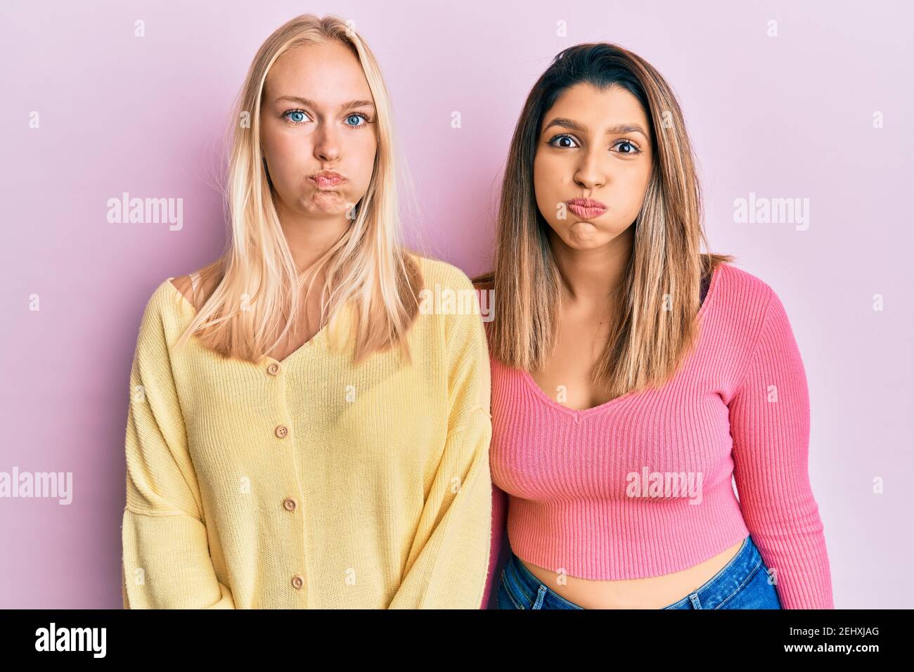 Two friends standing together over pink background puffing cheeks with ...