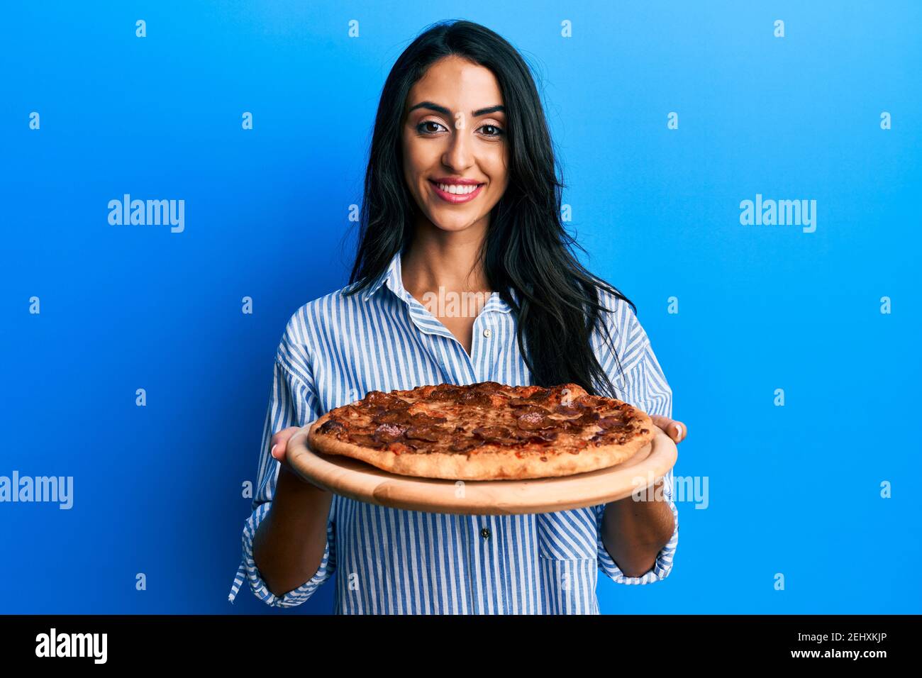 Beautiful hispanic woman holding tasty pepperoni pizza smiling with a ...