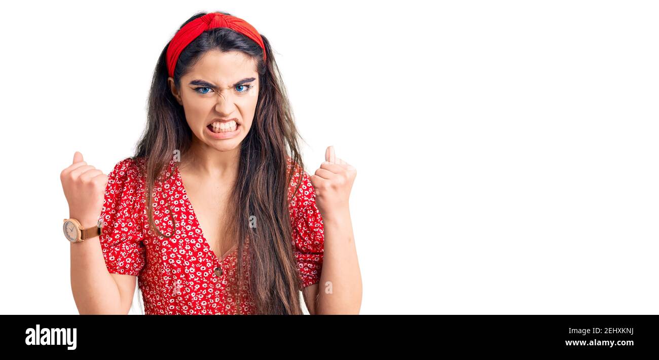 Brunette teenager girl wearing summer dress angry and mad raising fists ...