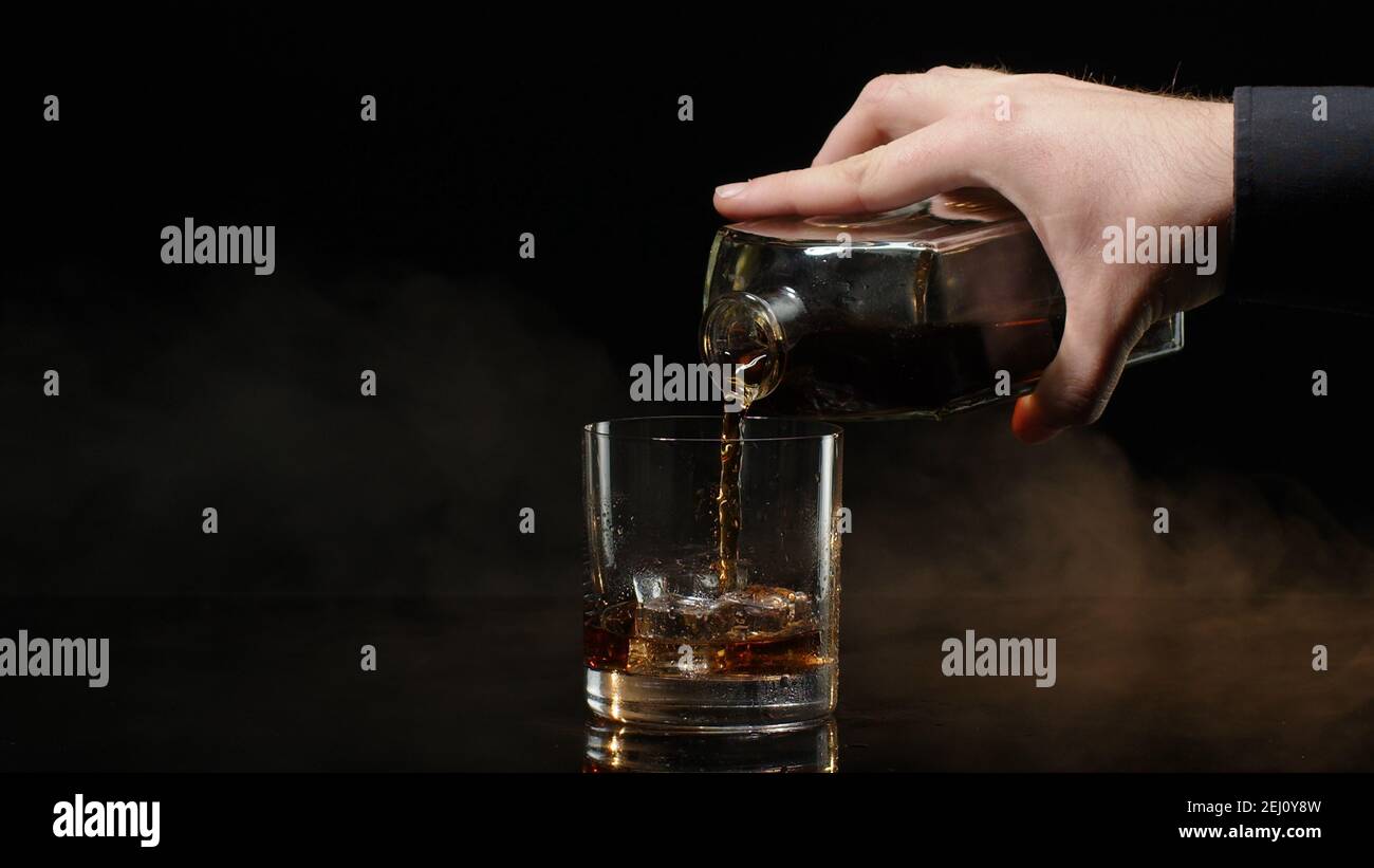 Barman pour golden whiskey, cognac, brandy from bottle into drinking glass with ice cubes on ...