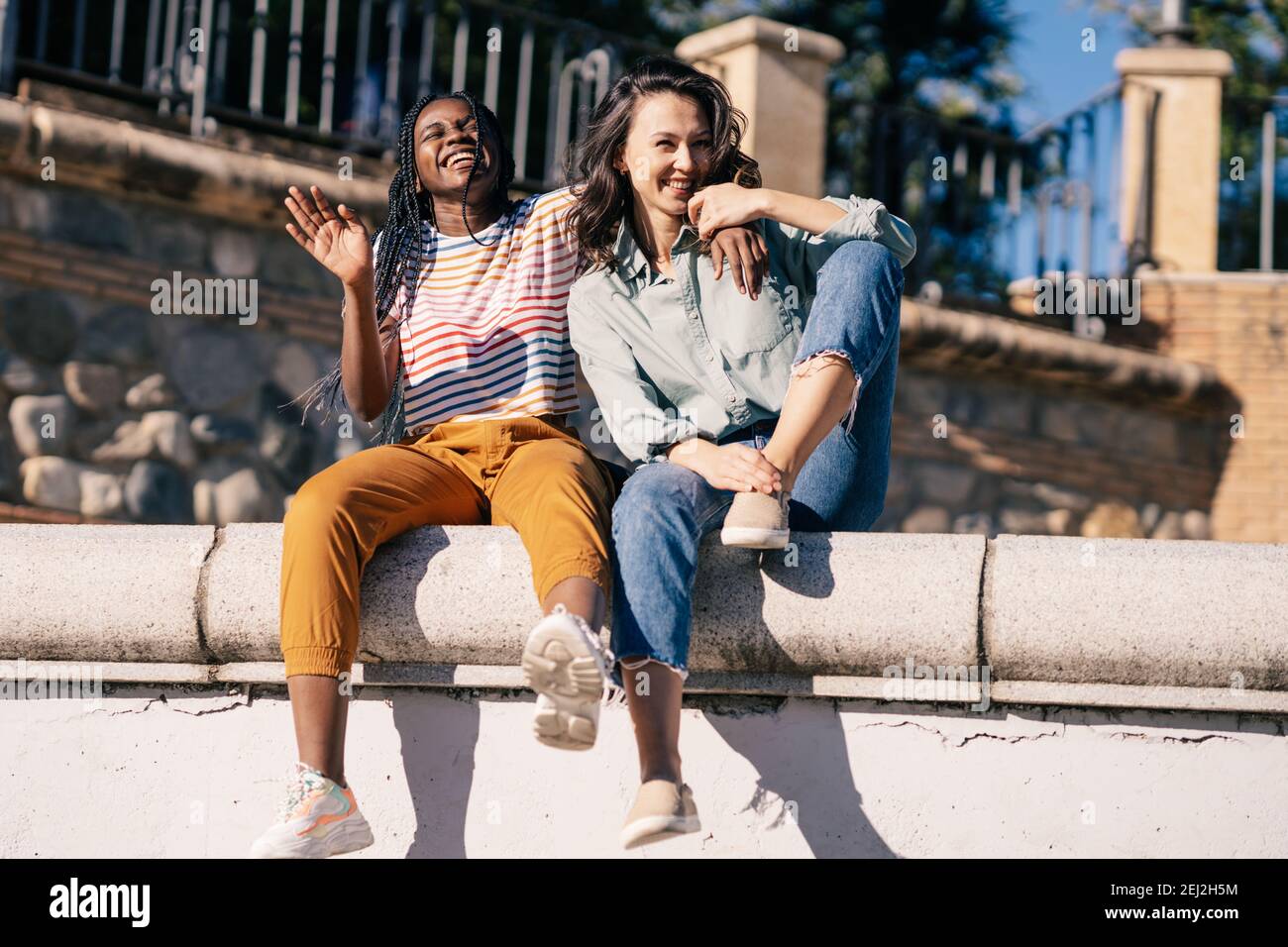 Two friends having fun together on the street sitting on a urban wall ...