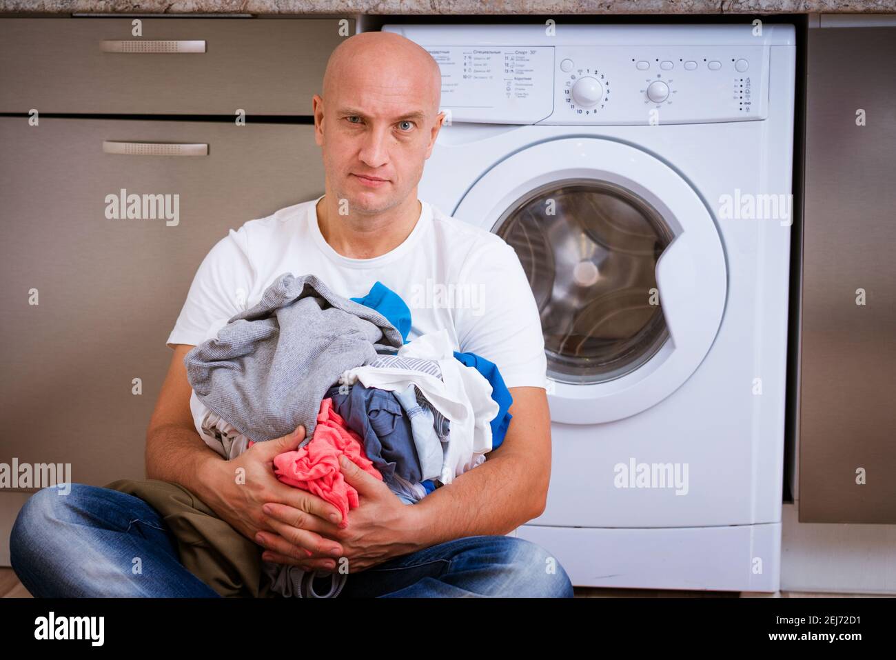 Bald man tired sitting near the washing machine holding dirty laundry ...