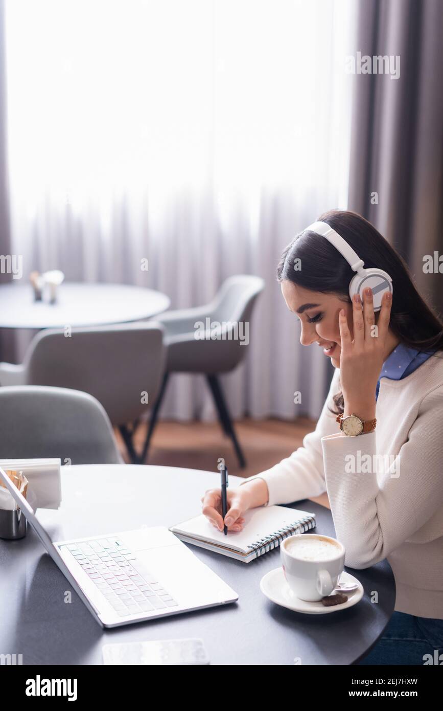 positive freelancer touching wireless headphones while writing in ...