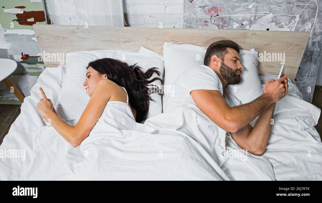 young couple messaging on smartphones while lying in bed back to back ...