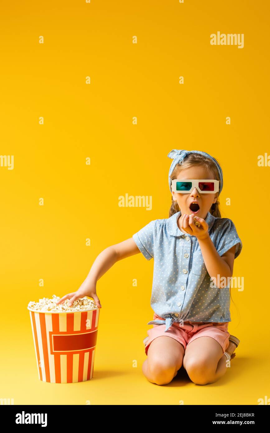 amazed kid in 3d glasses eating popcorn and reaching bucket on yellow ...