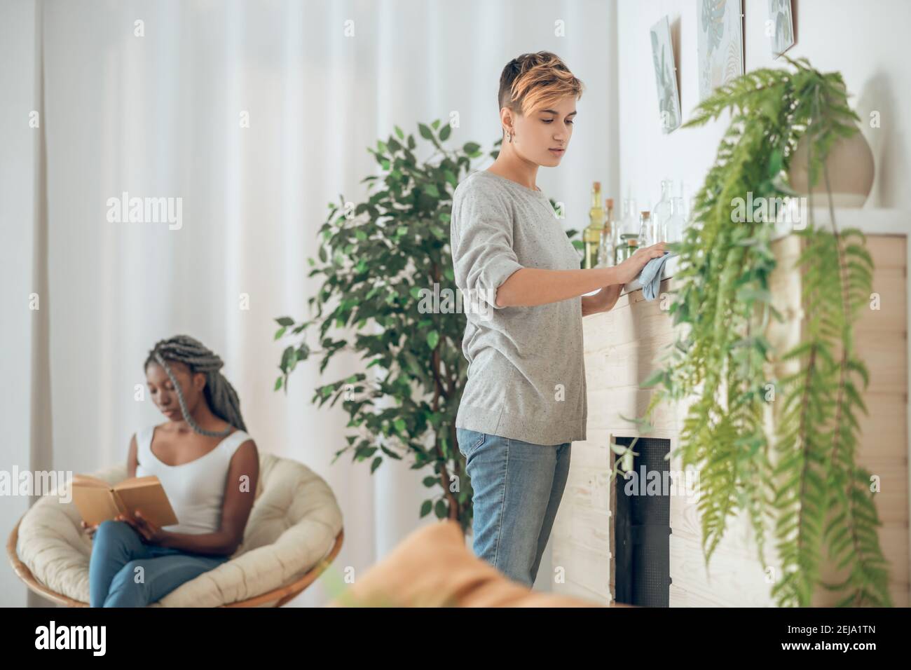 Fair-haired girl cleaning the room, her girlfriend sitting in the chair ...