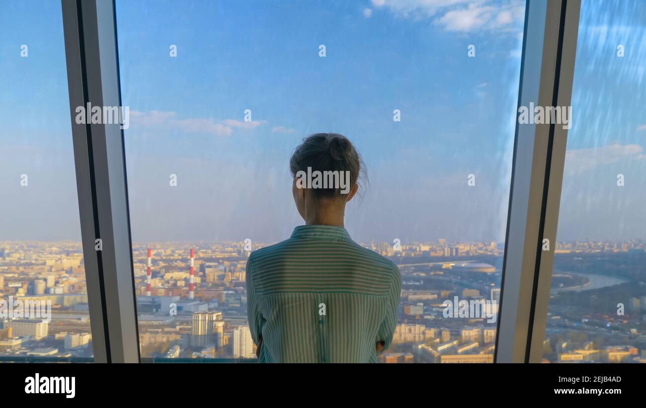 Pensive woman looking at cityscape through window of skyscraper - back ...