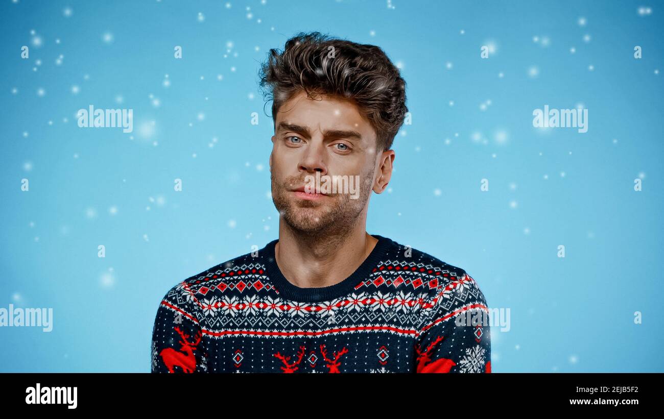 Young man in sweater looking at camera under falling snow on blue ...