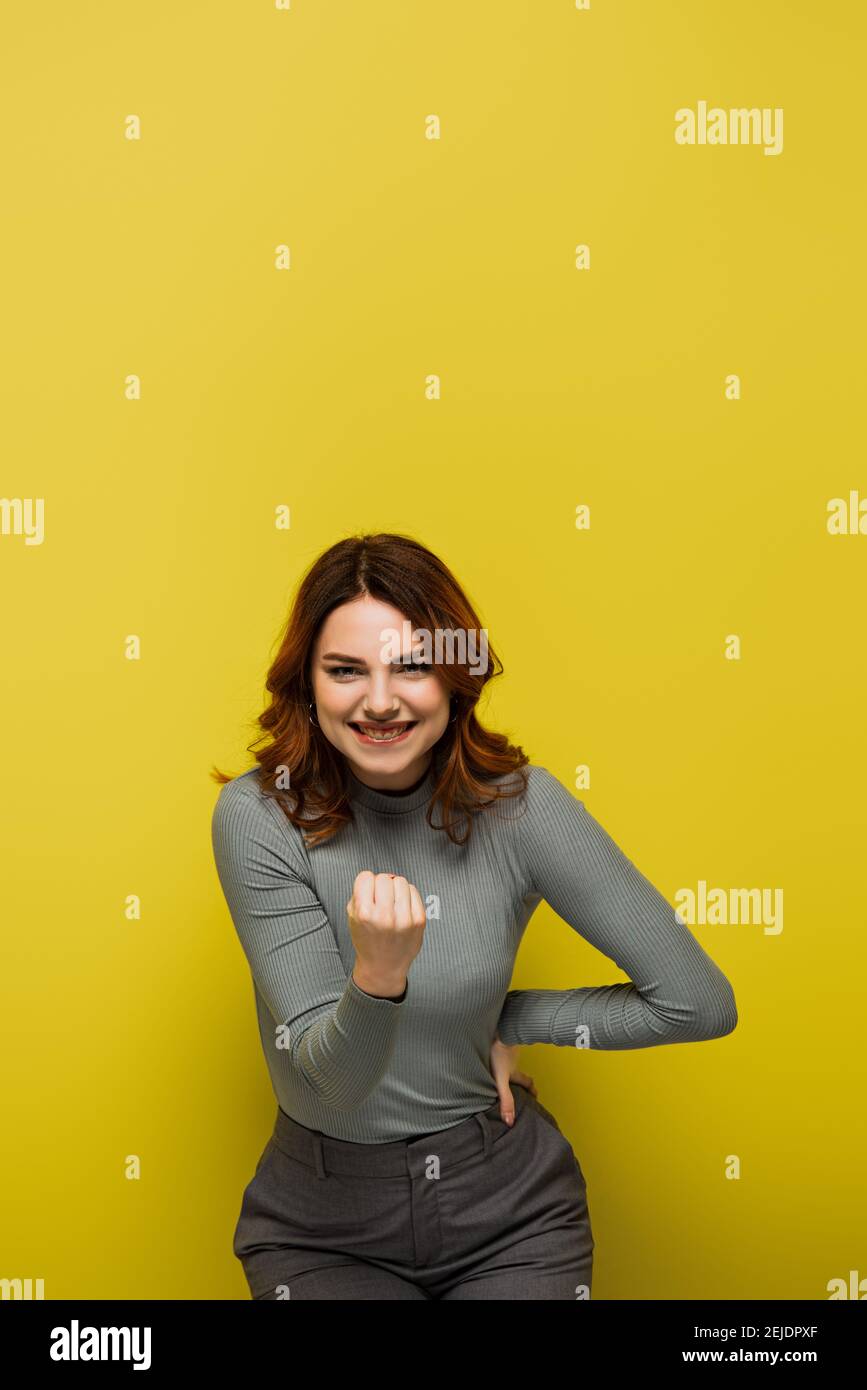 curly woman with clenched fist and hand on hip standing and smiling on ...