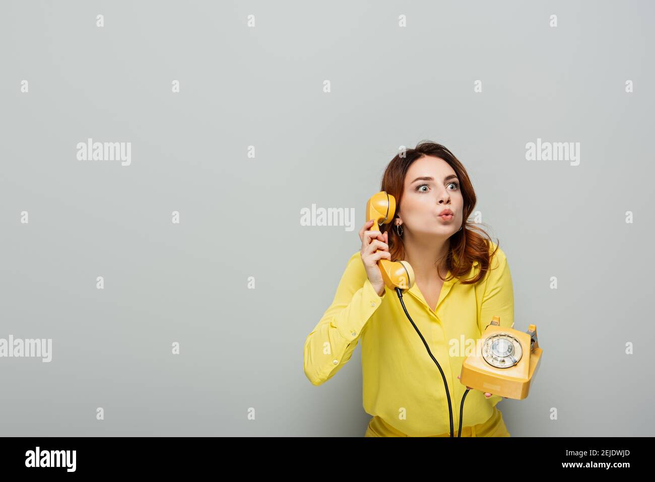 surprised woman pouting lips while talking on vintage telephone on grey ...