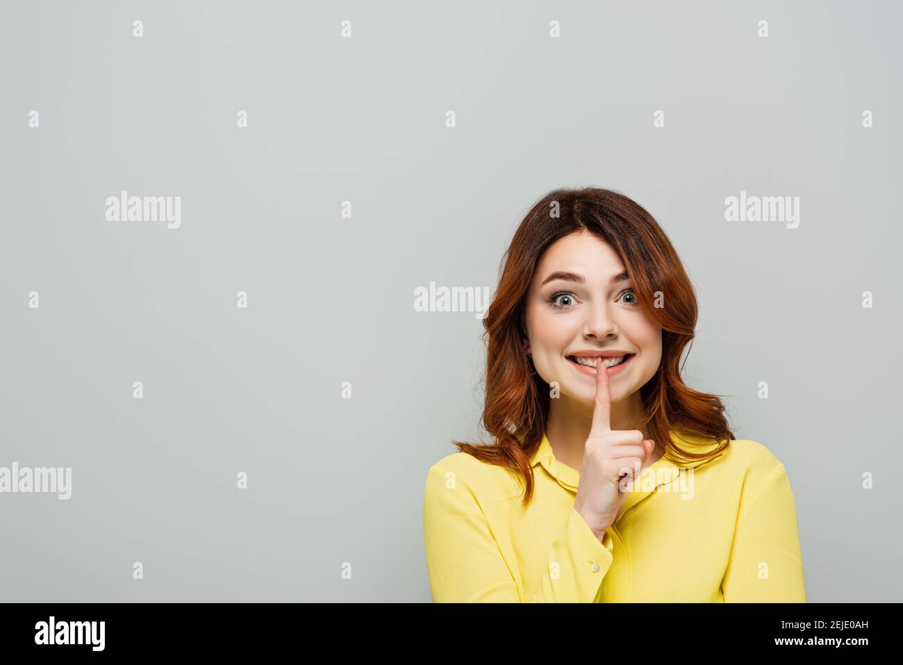smiling woman with curly hair showing hush gesture isolated on grey ...