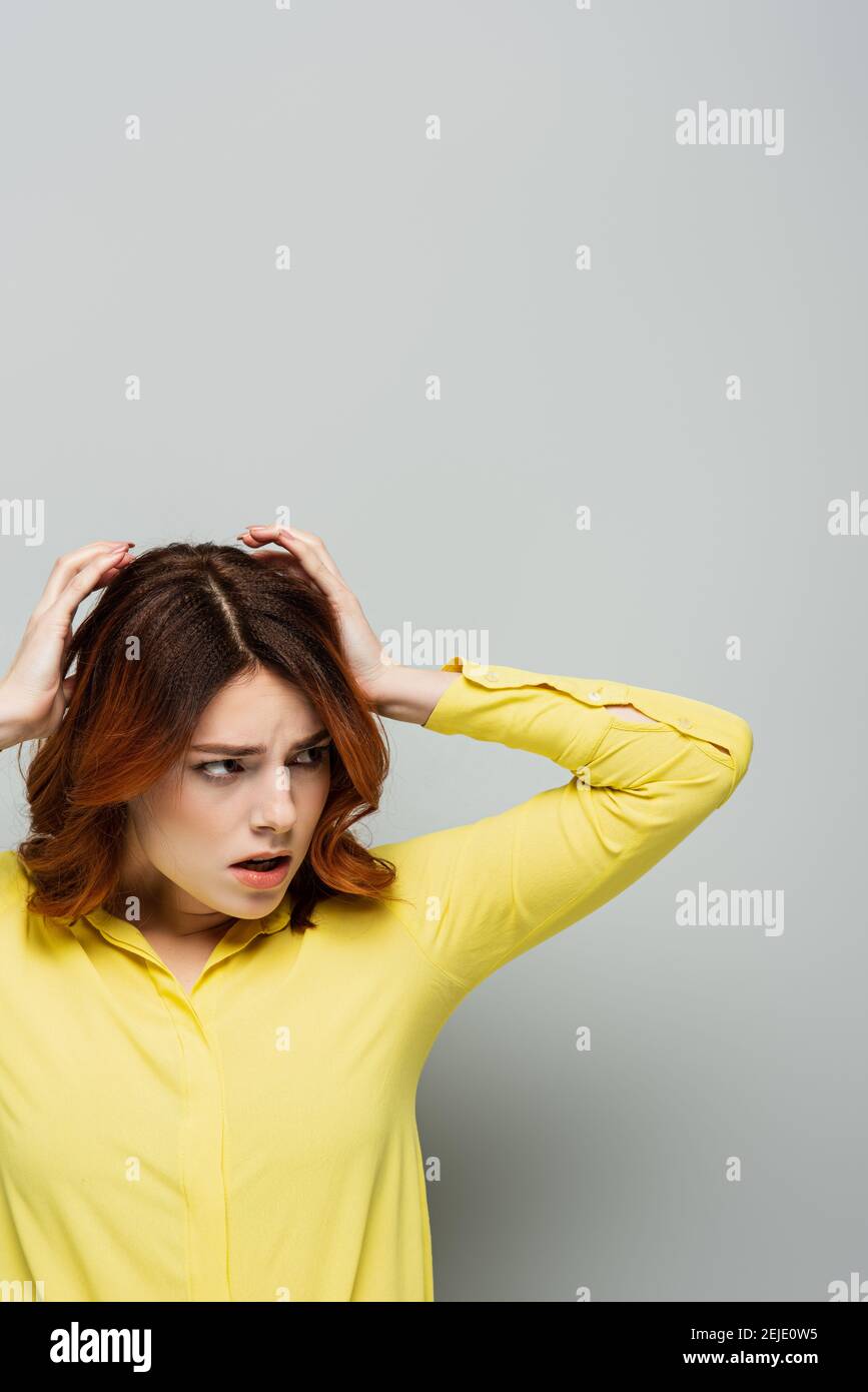 tense woman with curly hair touching head while looking away on grey ...