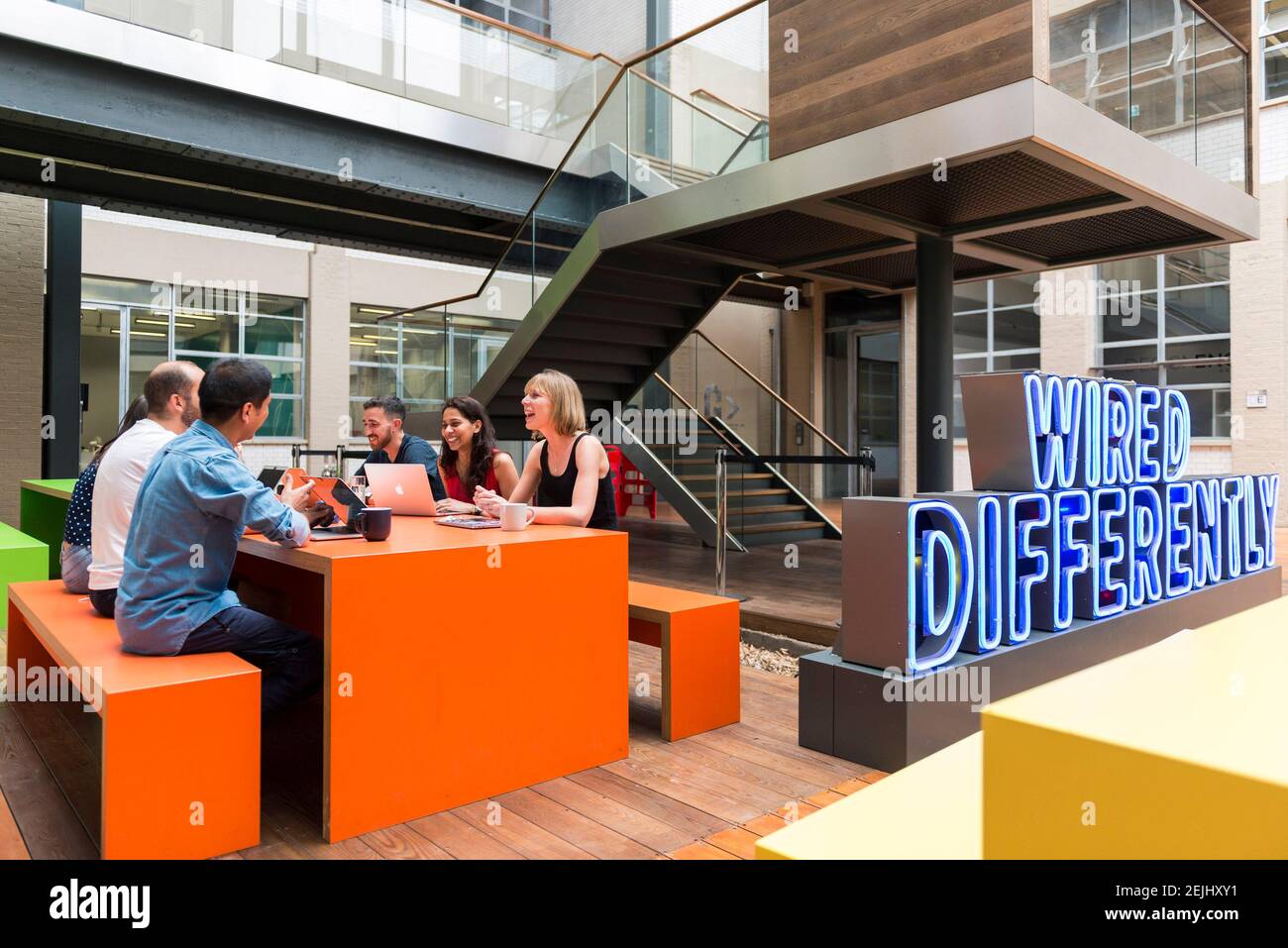 Six workers sit round an open plan meeting table in a communal space in ...
