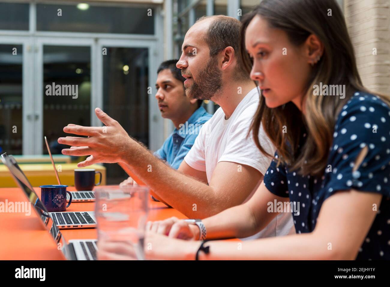 Six workers sit round an open plan meeting table in a communal space in ...