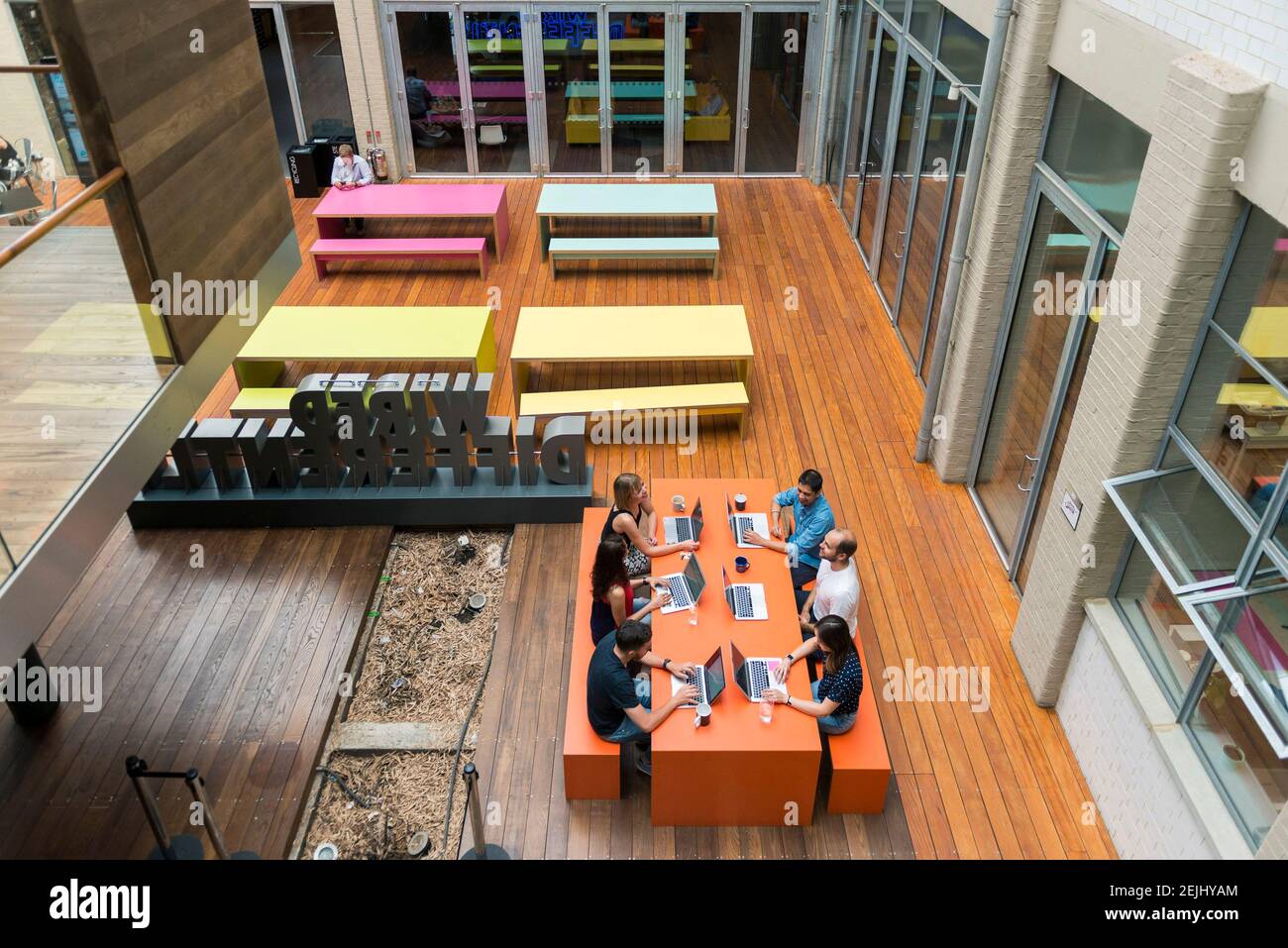 Six workers sit round an open plan meeting table in a communal space in ...
