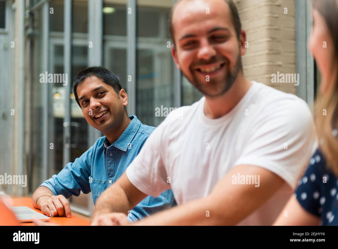 Six workers sit round an open plan meeting table in a communal space in a modern and bright ...
