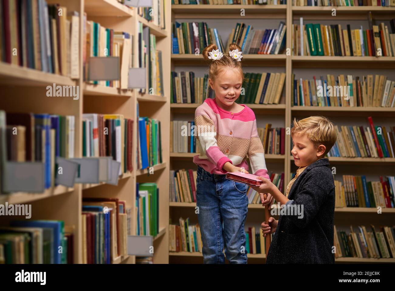 two school children help each other to get a book from shelf, stand ...