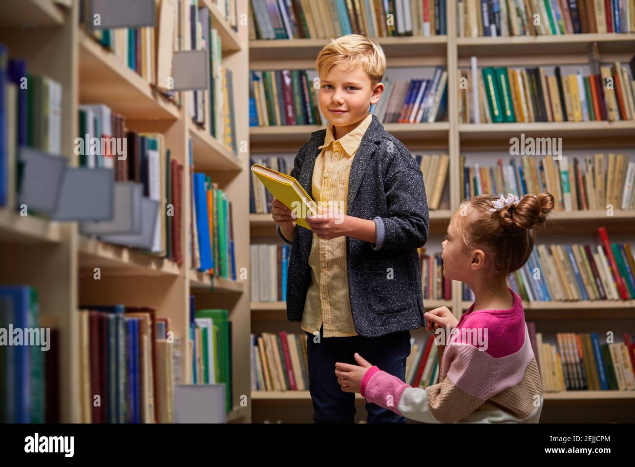 two school children help each other to get a book from shelf, stand ...
