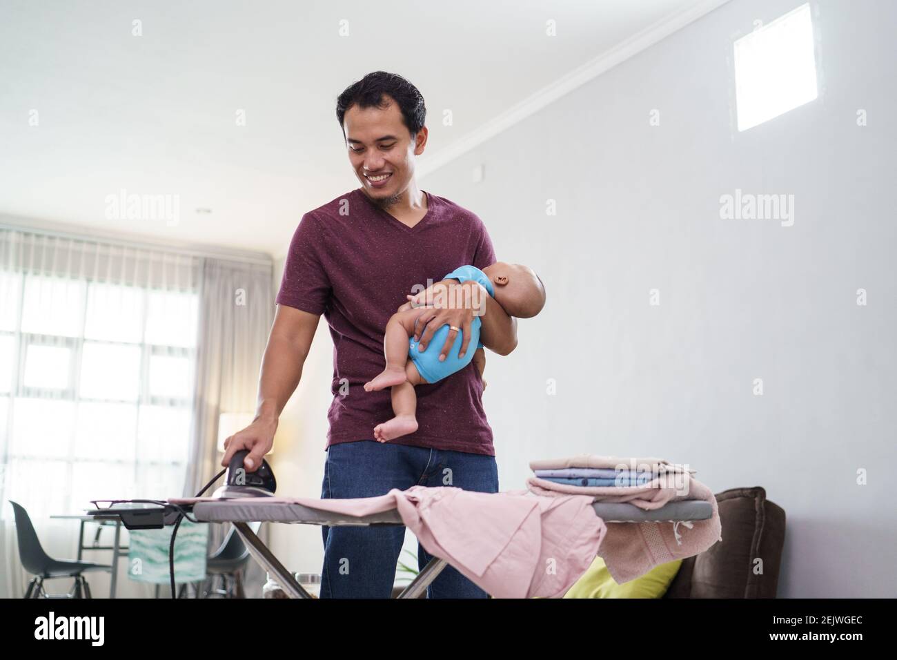 portrait of asian father ironing his clothes while holding his infant ...