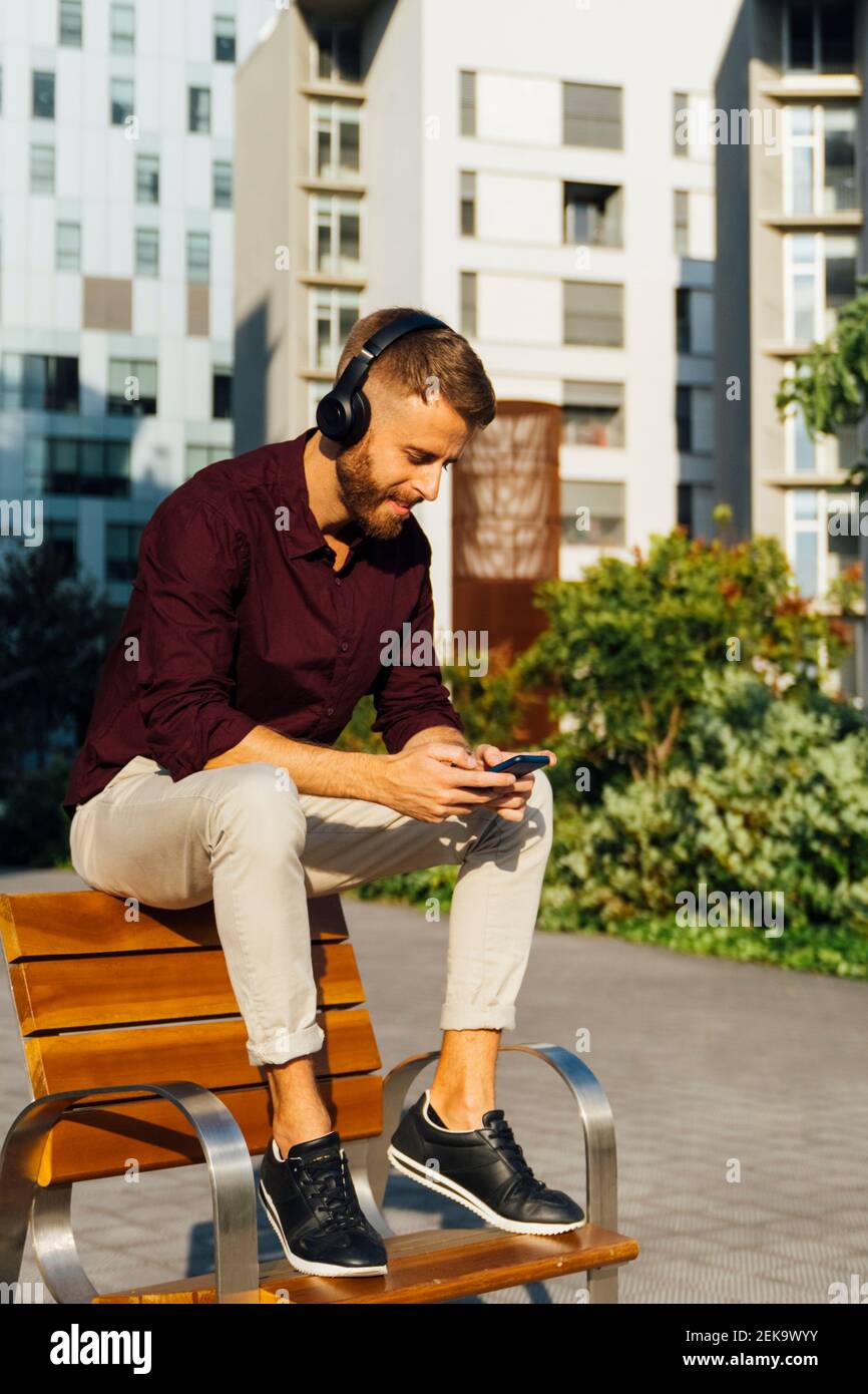 Businessman wearing headphones using smart phone while sitting on bench ...