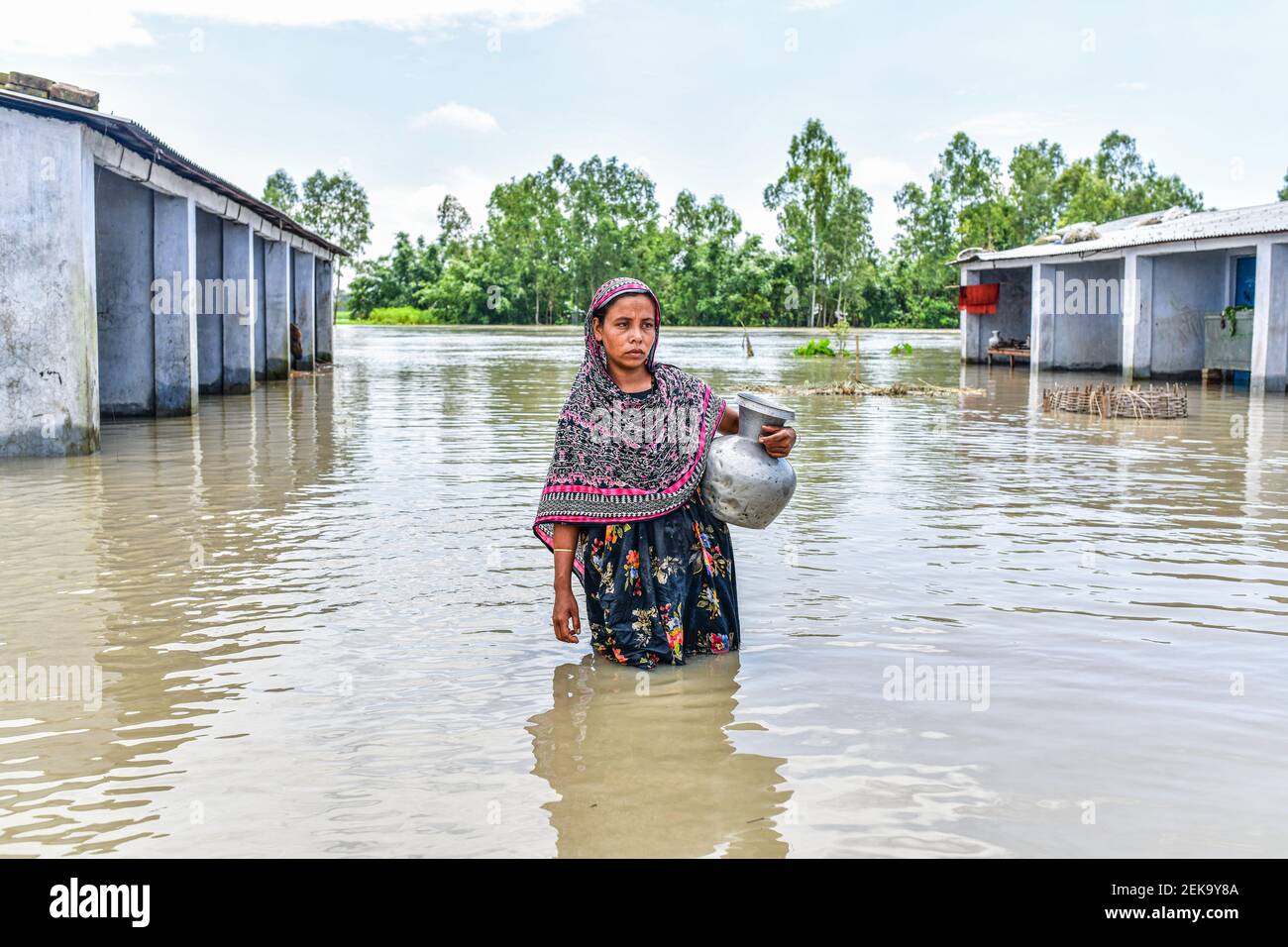 A woman walks through floodwaters carrying a jar to collect water. At ...