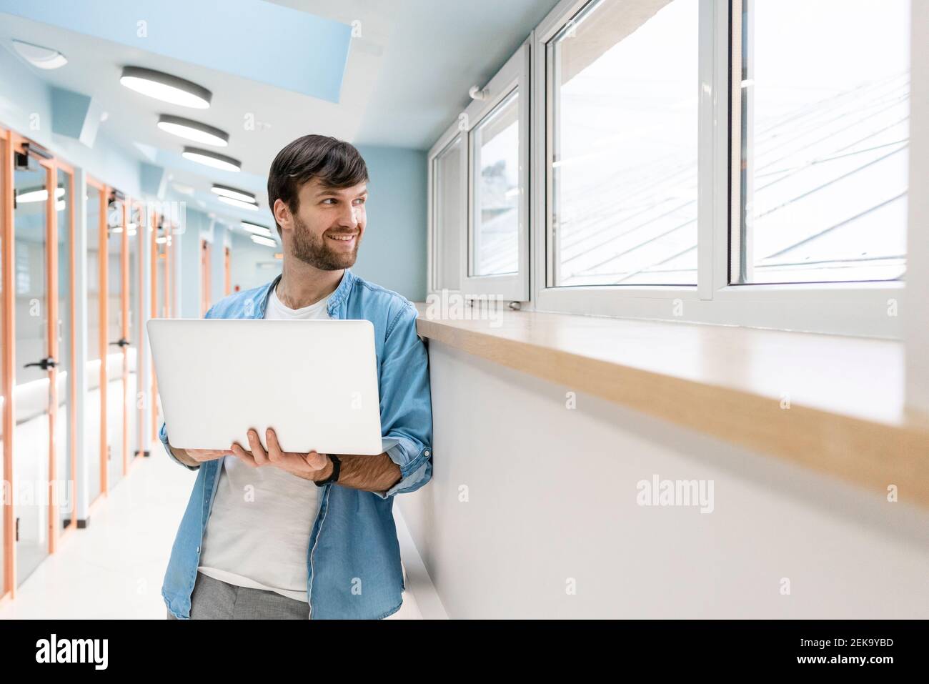 Smiling businessman looking through window while holding laptop in ...