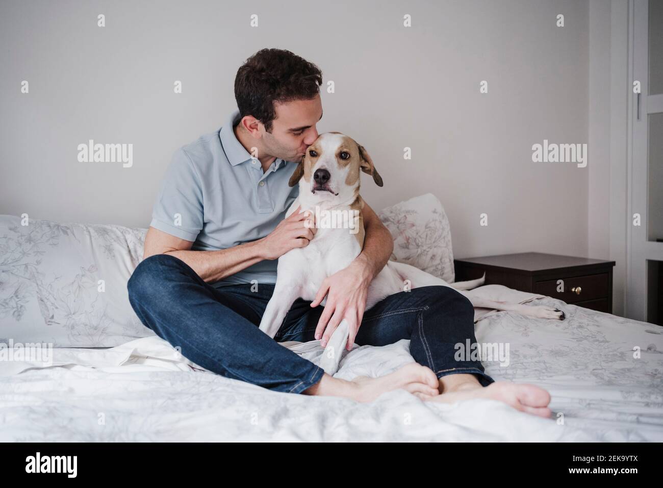 Mid adult man kissing dog while sitting on bed against wall at home ...