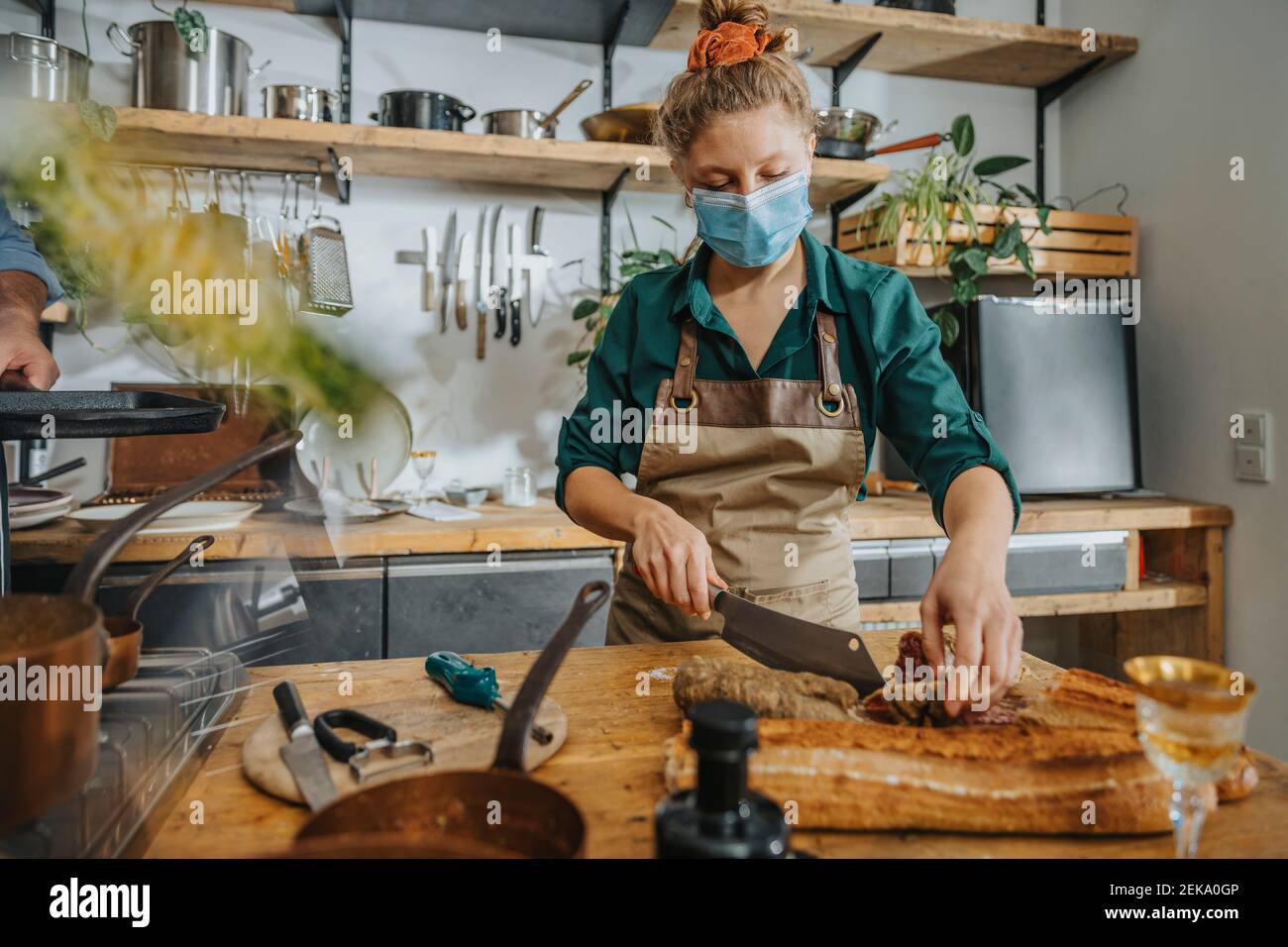 Young chef wearing protective face mask cutting salami while standing ...