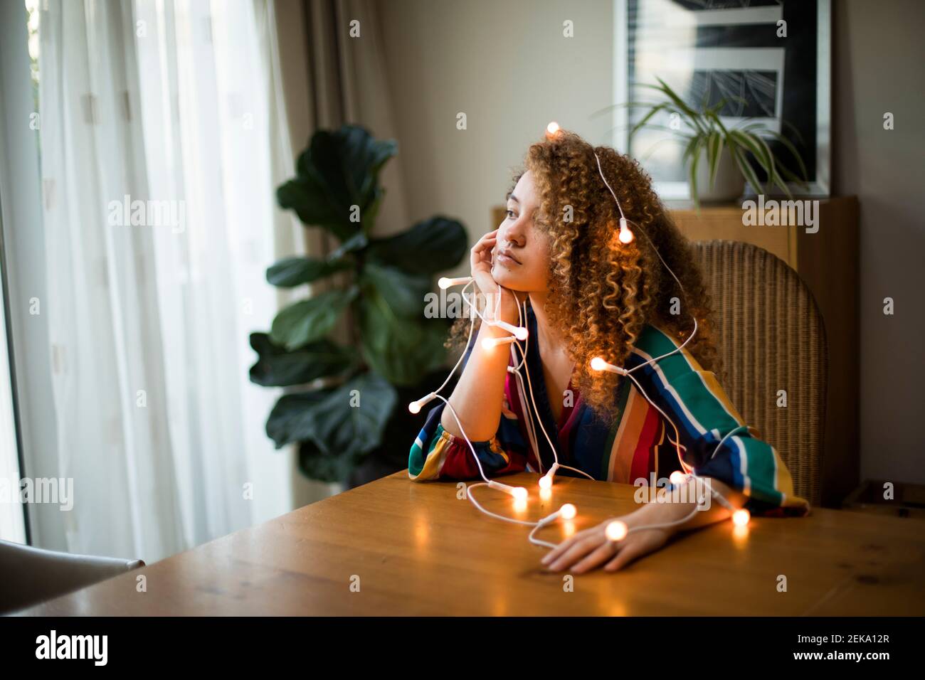 Thoughtful young woman with Christmas lights sitting on chair at home ...