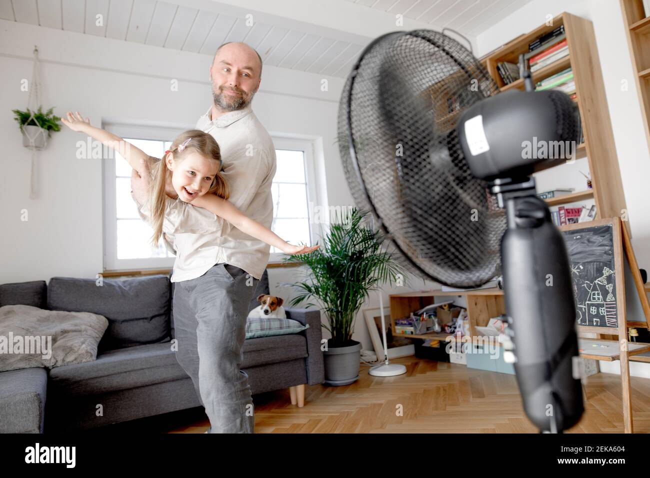 Father playing with cheerful daughter in front of electric fan at home ...