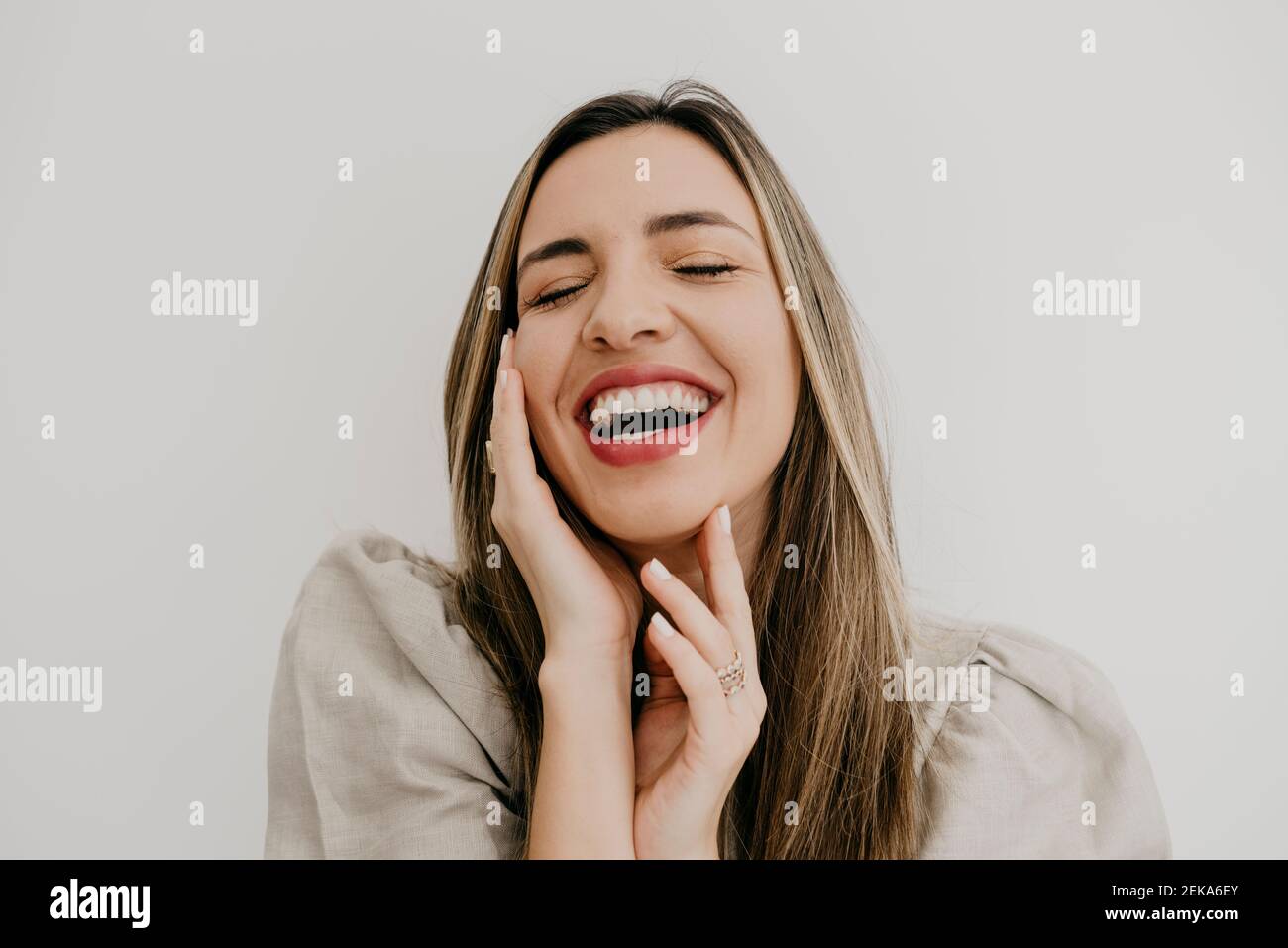 Laughing woman touching cheek with eyes closed against white background ...