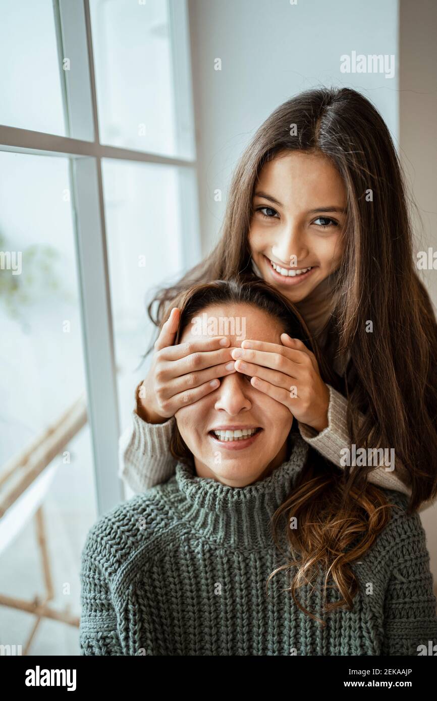 Happy daughter covering eyes of mother while sitting by window at home ...