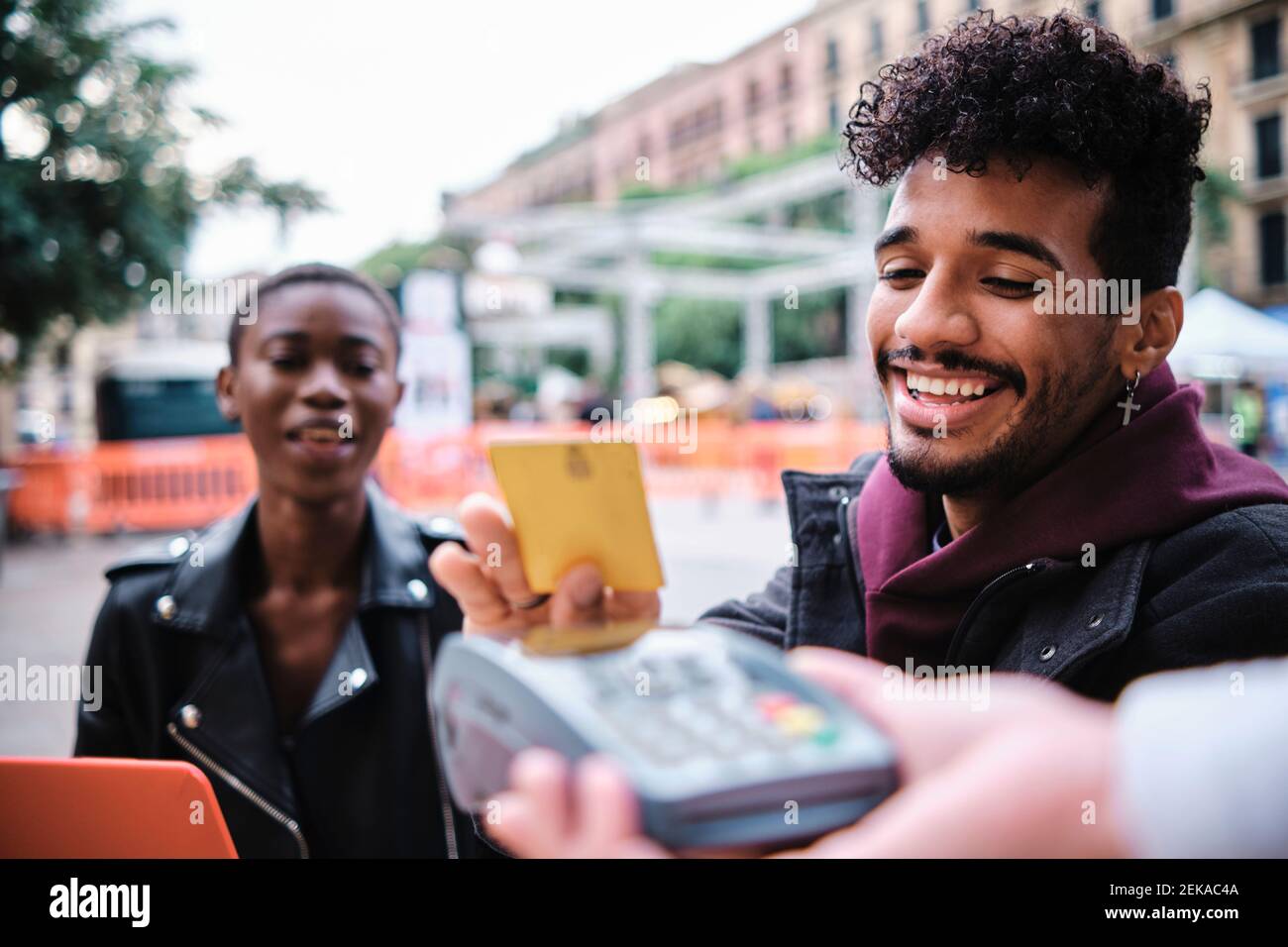 Smiling man paying through card whiles sitting with friend at sidewalk ...