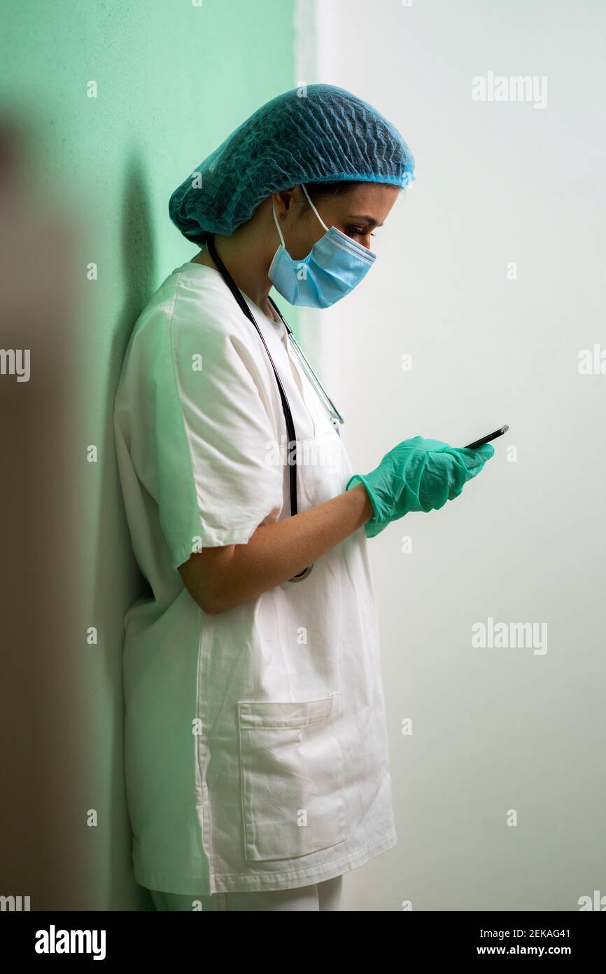 Female doctor using mobile phone while standing against wall at clinic ...