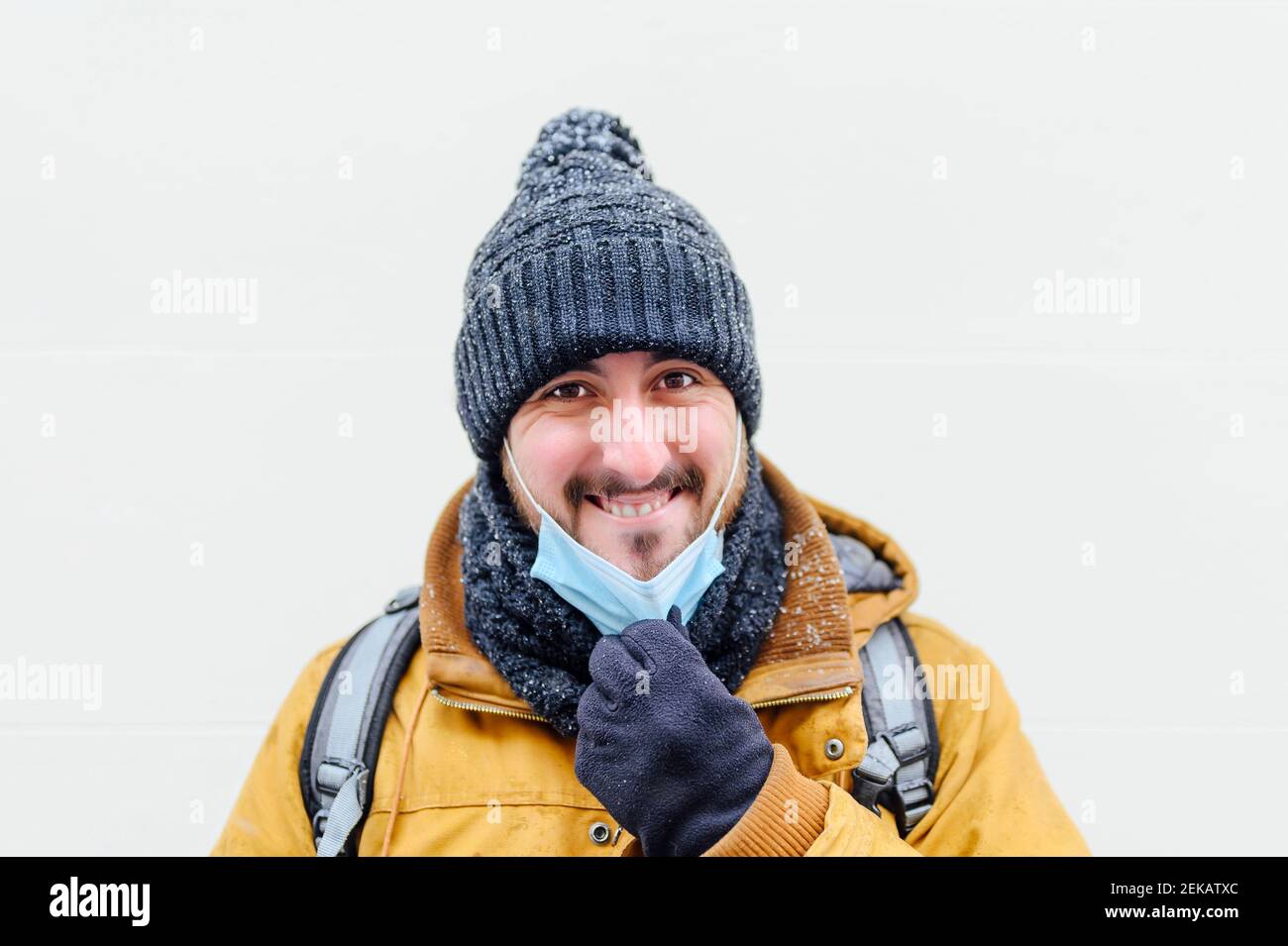 Smiling man pulling mask against white wall during COVID-19 Stock Photo ...